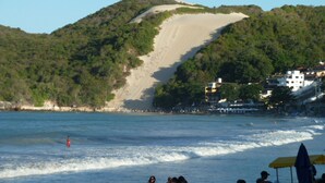 Uma praia com pessoas, uma duna de areia e edifícios ao fundo.