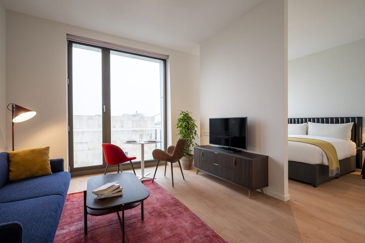 Quarto de hotel moderno com sofá, mesa de jantar, cadeira vermelha, televisão, cama com cabeceira e uma janela grande.