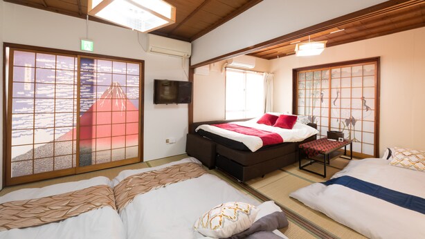 Um quarto tradicional japonês com tatames, uma cama com roupa de cama vermelha, uma pequena mesa e portas deslizantes com vista para uma montanha.