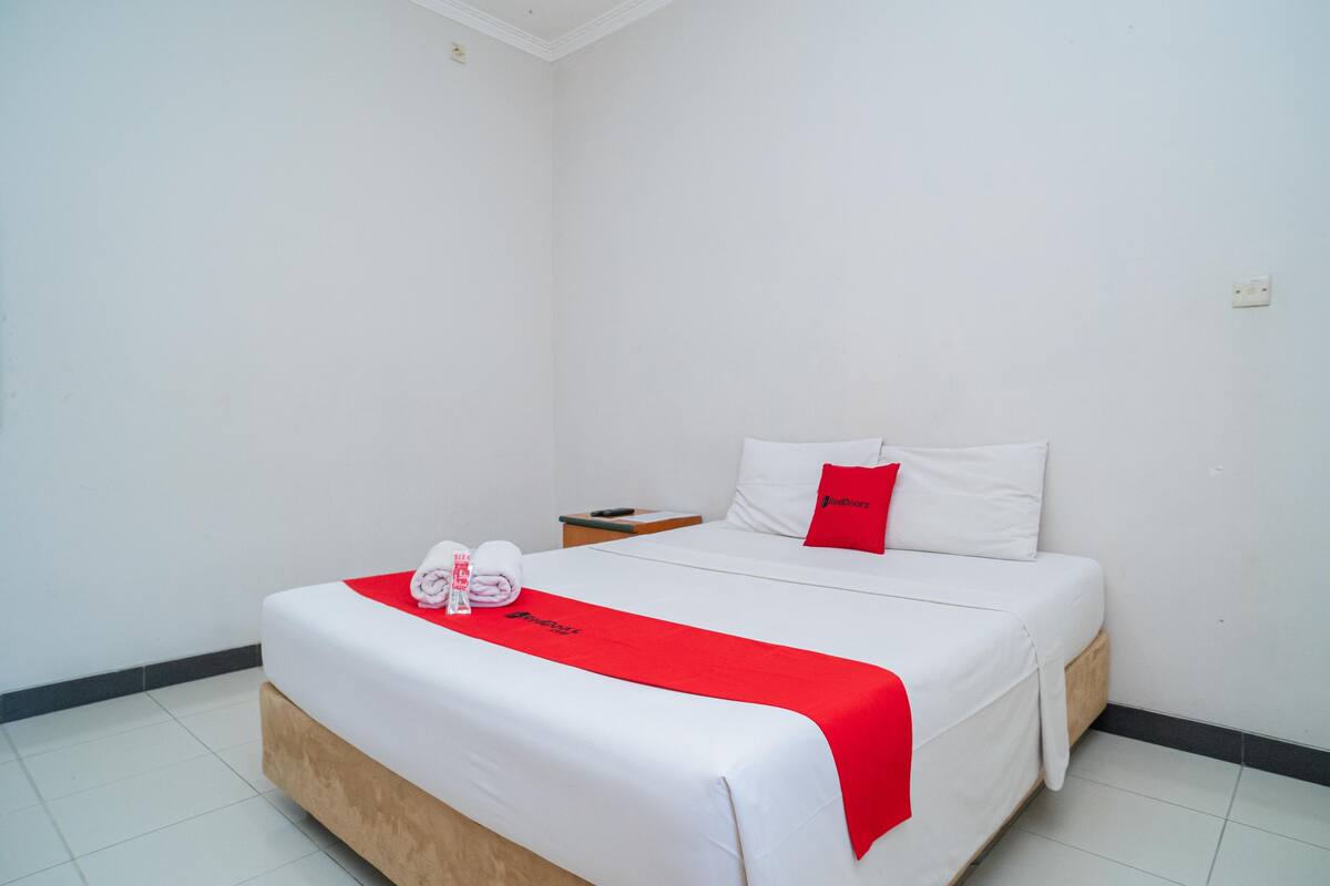 Cama bem arrumada com uma toalha vermelha e um travesseiro vermelho, uma mesa de cabeceira de madeira e uma parede branca.