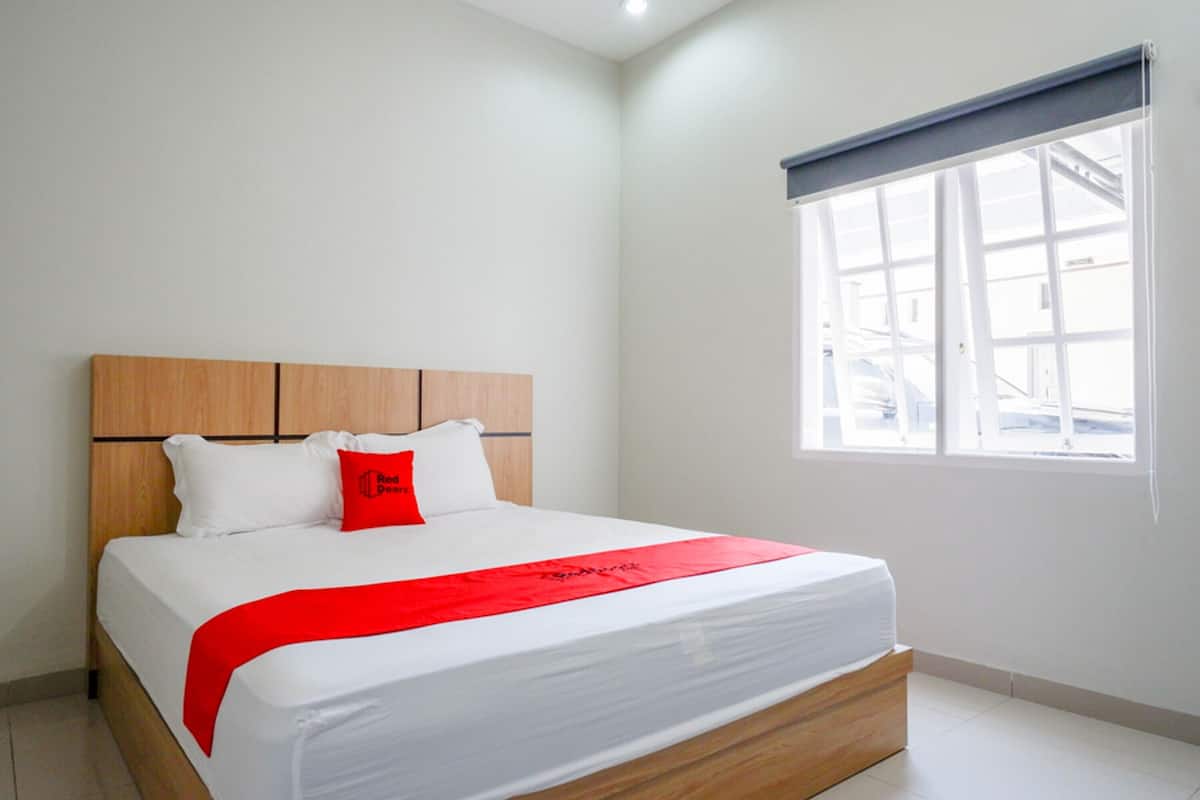 Uma cama bem arrumada com cabeceira de madeira, roupa de cama branca e uma almofada vermelha decorativa.