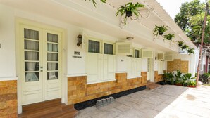 Entrada de hotel com varanda de azulejos, degraus que levam à porta e plantas em vasos penduradas acima.