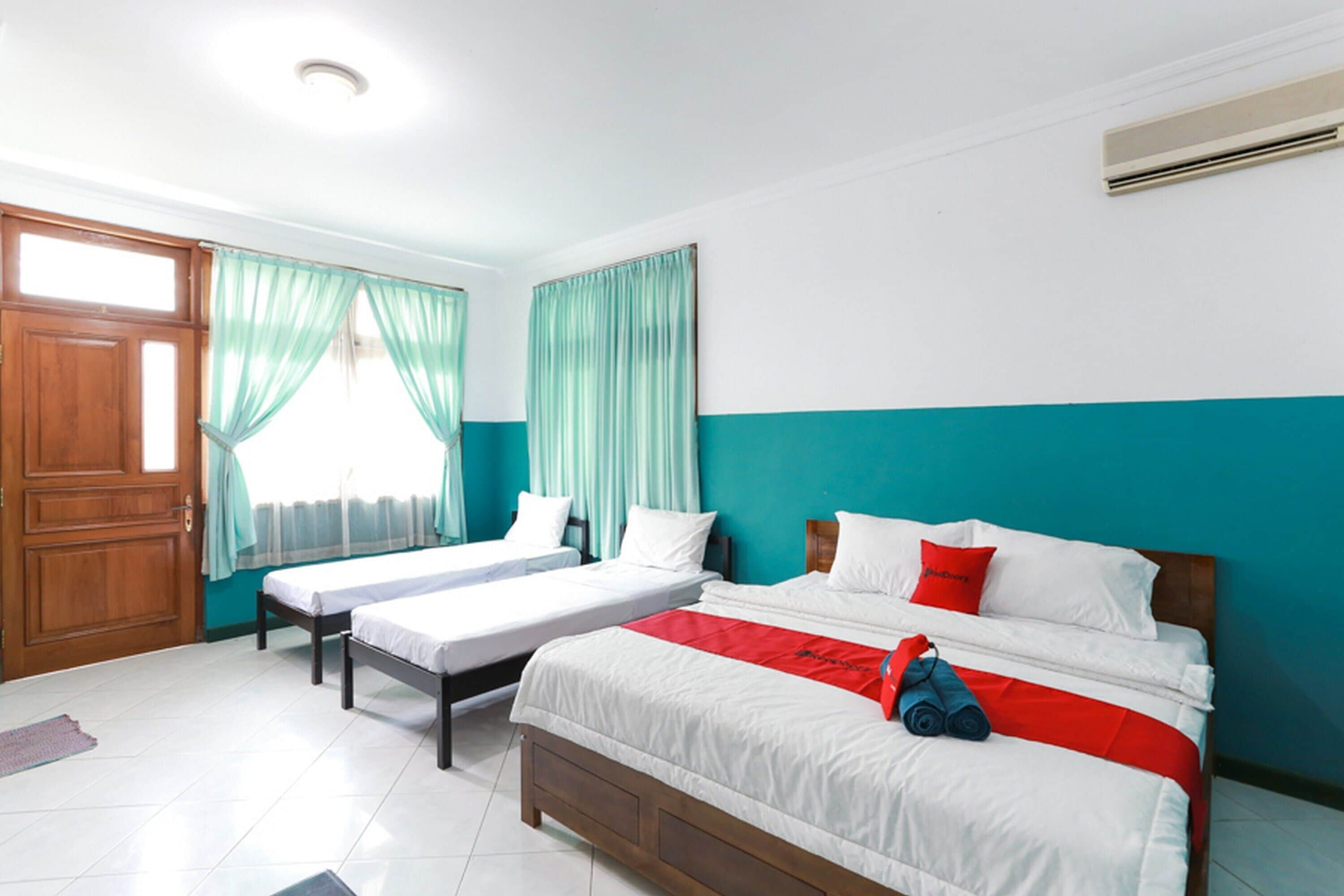 Quarto de hotel com duas camas de solteiro, guarda-roupa de madeira, janela com cortinas e uma parede com detalhes em verde-água.