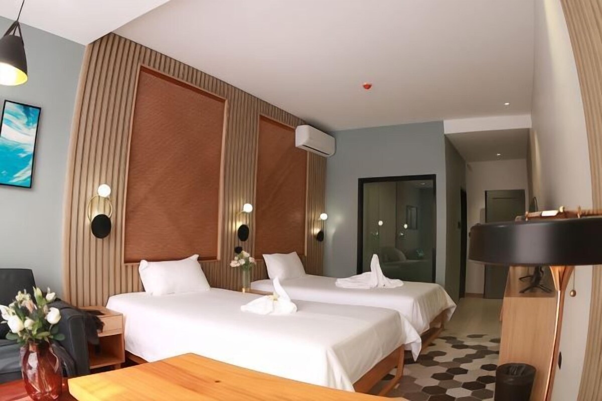 Quarto de hotel com duas camas, uma escrivaninha de madeira, um vaso com flores e um ar condicionado fixado na parede.