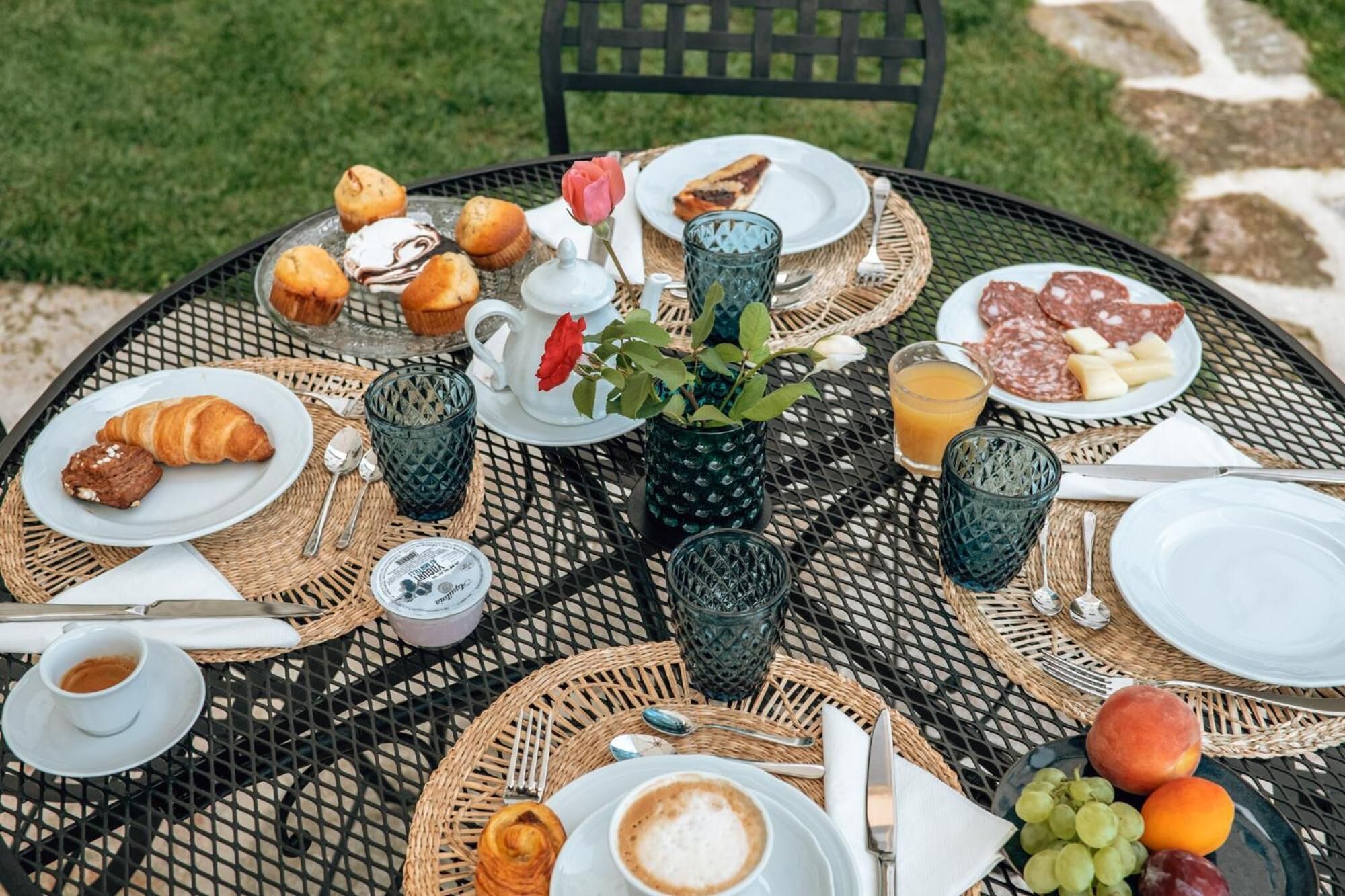 Mesa de jantar redonda ao ar livre, com diversas opções de café da manhã, incluindo doces, frutas e bebidas, sobre uma mesa de metal com jogos americanos trançados.