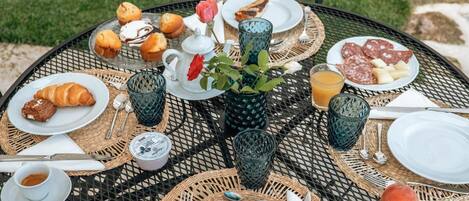 Mesa de jantar redonda ao ar livre, com diversas opções de café da manhã, incluindo doces, frutas e bebidas, sobre uma mesa de metal com jogos americanos trançados.