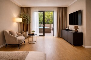 Quarto de hotel com cama, área de estar com uma cadeira e uma pequena mesa, televisão na parede e vista para área externa através de uma porta de vidro.