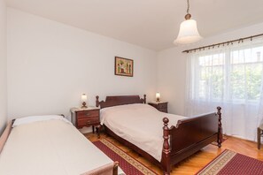 Quarto com duas camas de solteiro, cabeceiras de madeira, mesas de cabeceira com abajures, luminária pendente, janela com cortinas leves e um quadro na parede.
