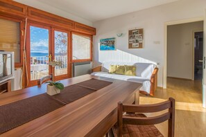 Quarto com mesa de jantar de madeira, sofá com almofadas, televisão e vista para o mar através de grandes janelas.