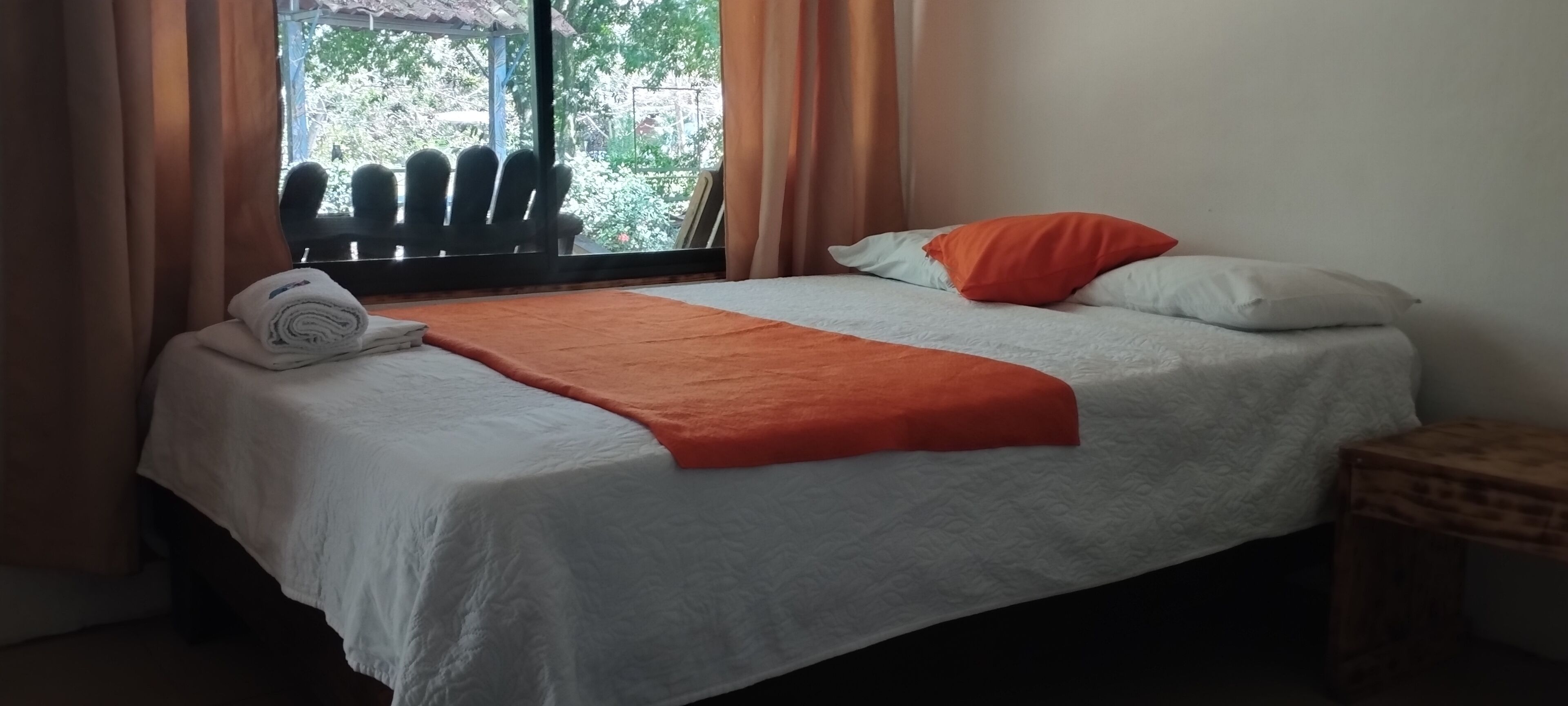 Uma cama bem arrumada com roupa de cama branca e um cobertor laranja, uma mesa de cabeceira de madeira e uma janela com cortinas.