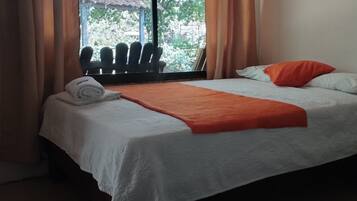Uma cama bem arrumada com roupa de cama branca e um cobertor laranja, uma mesa de cabeceira de madeira e uma janela com cortinas.