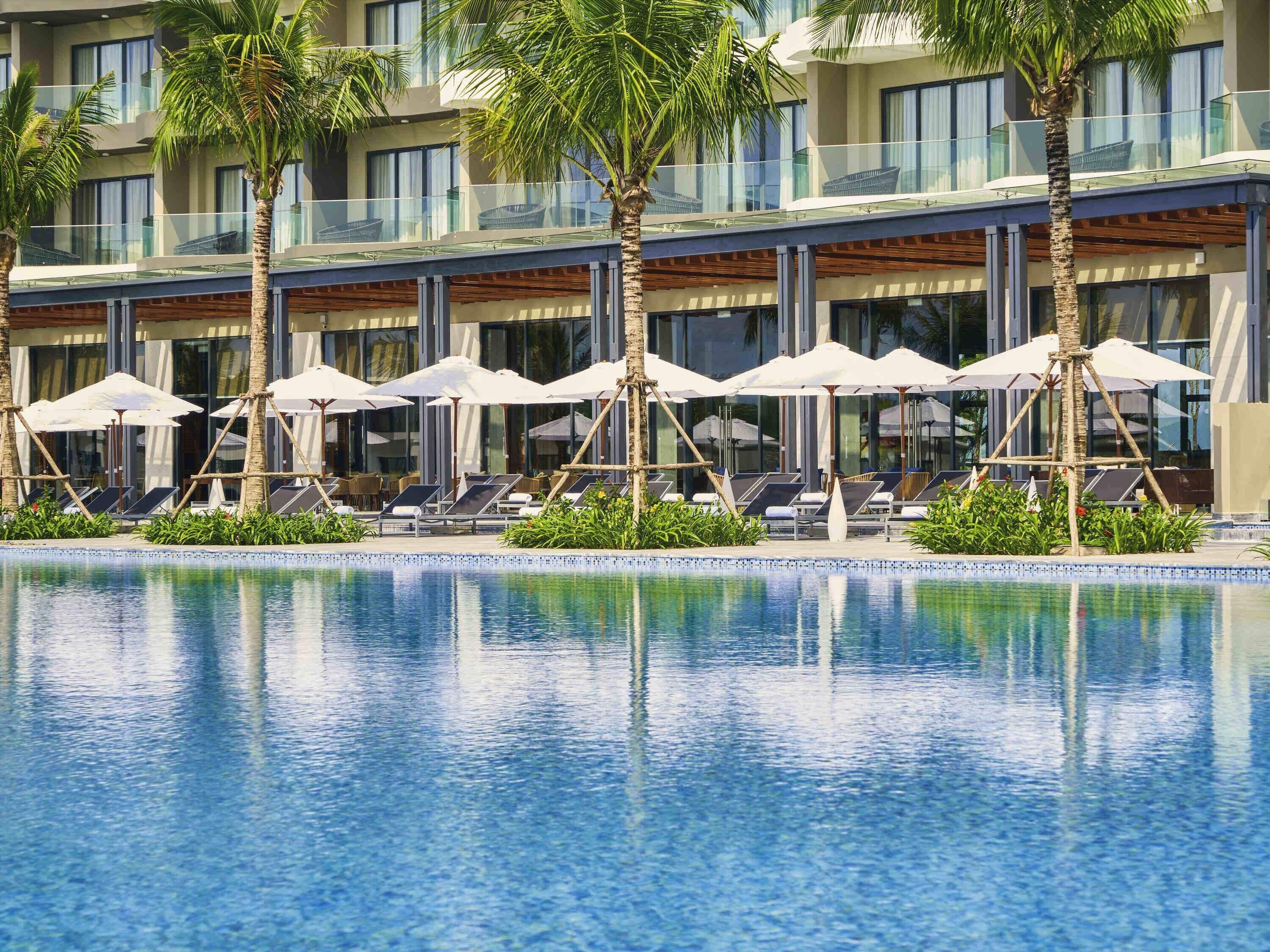 Um hotel com piscina, espreguiçadeiras, guarda-sóis e palmeiras.