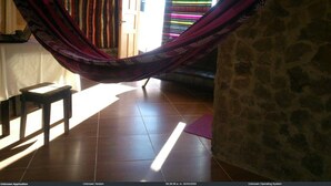 Quarto com rede, vigas de madeira, parede de pedra e vista para cortinas coloridas.
