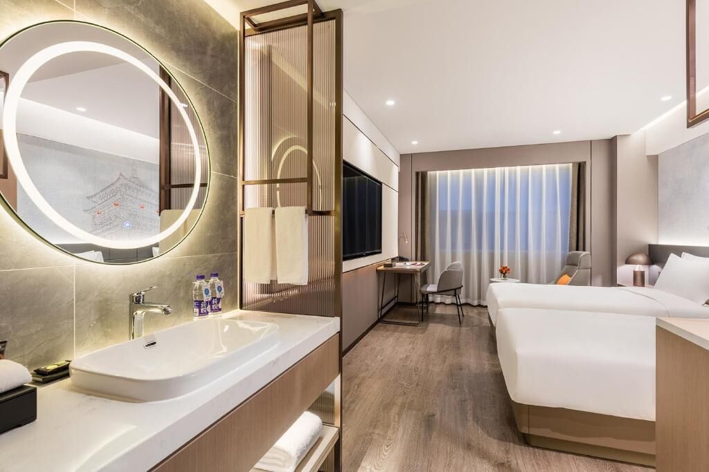 Quarto de hotel moderno com um espelho grande e redondo, pia, cama, escrivaninha e uma cadeira.