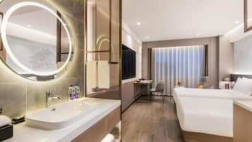 Quarto de hotel moderno com um espelho grande e redondo, pia, cama, escrivaninha e uma cadeira.