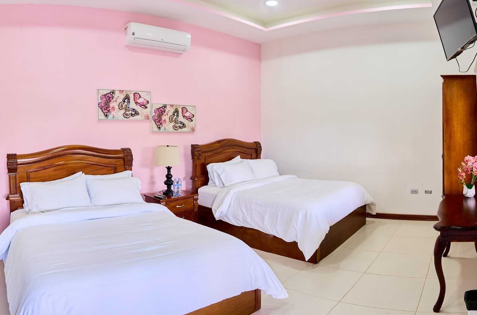 Quarto de hotel com duas camas de madeira, uma mesa de cabeceira com abajur e uma TV sobre suporte de madeira.
