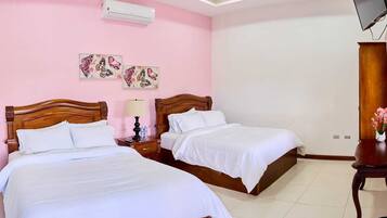 Quarto de hotel com duas camas de madeira, uma mesa de cabeceira com abajur e uma TV sobre suporte de madeira.