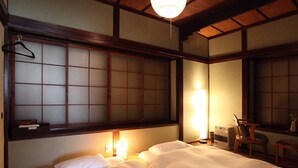 Quarto tradicional em estilo japonês, com tatames, uma cama baixa com roupa de cama branca, um aquecedor e um candeeiro suspenso.