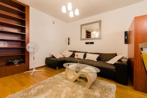 Uma sala de estar com sofá seccional, mesa de centro de vidro, ventilador, estante de livros e espelho na parede.