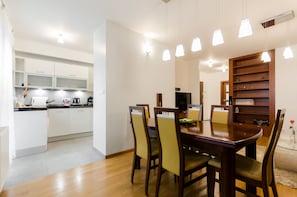 Uma zona de refeições moderna com uma mesa e cadeiras de madeira, uma cozinha ao fundo e iluminação pendente.