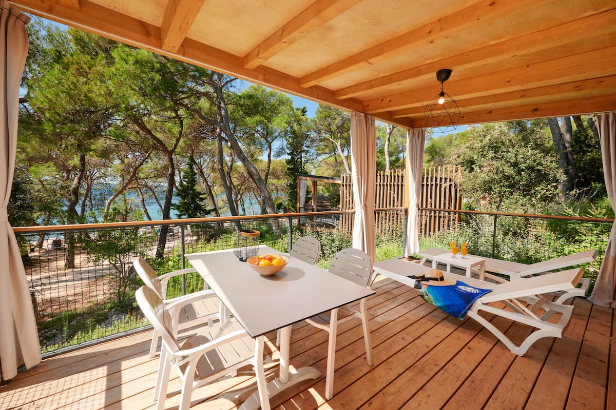 Um deck de madeira com uma mesa e cadeiras, com vista para uma área arborizada e o mar.