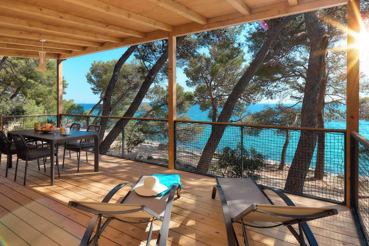 Um deck de madeira com mesa e cadeiras, com vista para uma praia, árvores e céu azul límpido.