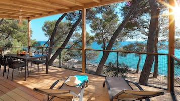 Um deck de madeira com mesa e cadeiras, com vista para uma praia, árvores e céu azul límpido.