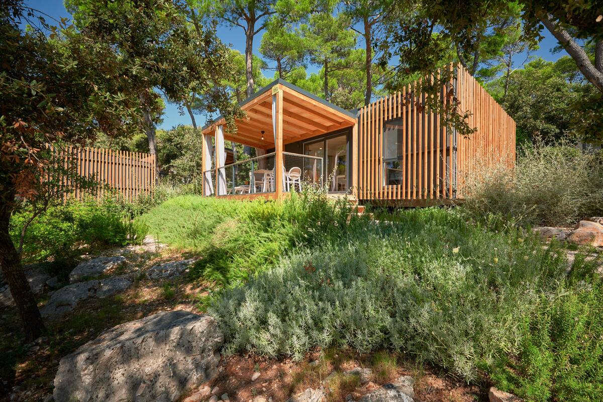Uma cabana moderna de madeira com varanda coberta e janelas amplas, rodeada por vegetação e árvores.