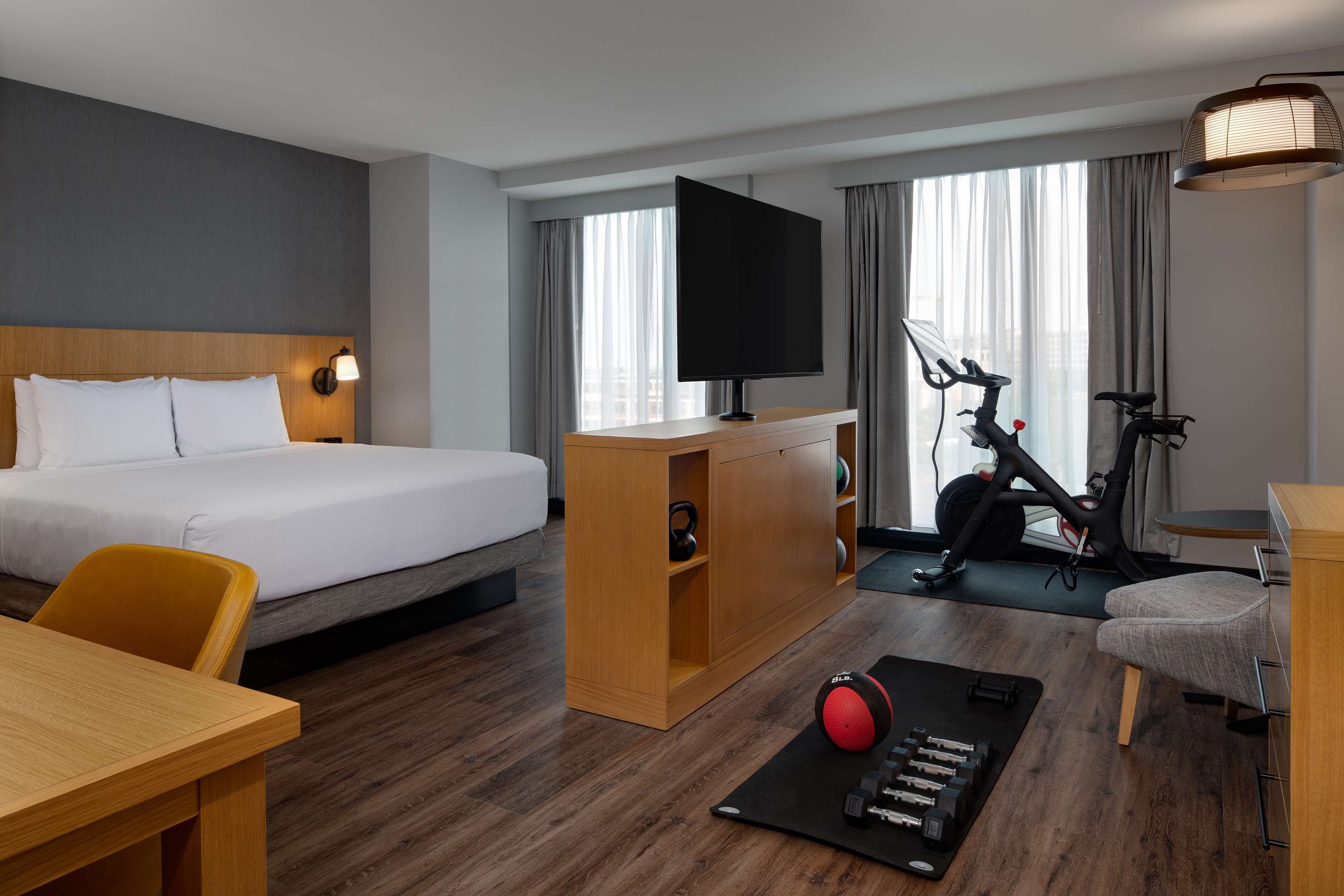 Quarto de hotel com cama, escrivaninha, televisão, bicicleta ergométrica e colchonete de ginástica.