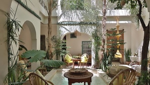 Um pátio interno com uma mesa e cadeiras no centro, rodeado por plantas grandes e uma fonte decorativa.