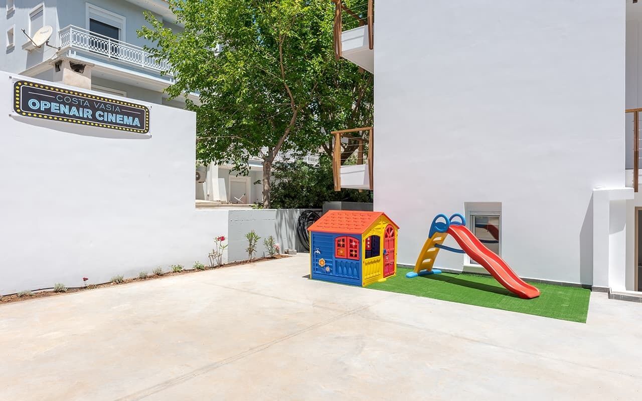 Uma área de recreação infantil com um escorrega e um pequeno parquinho, uma placa para 'Costa Vasia Openair Cinema' e um edifício com varandas.