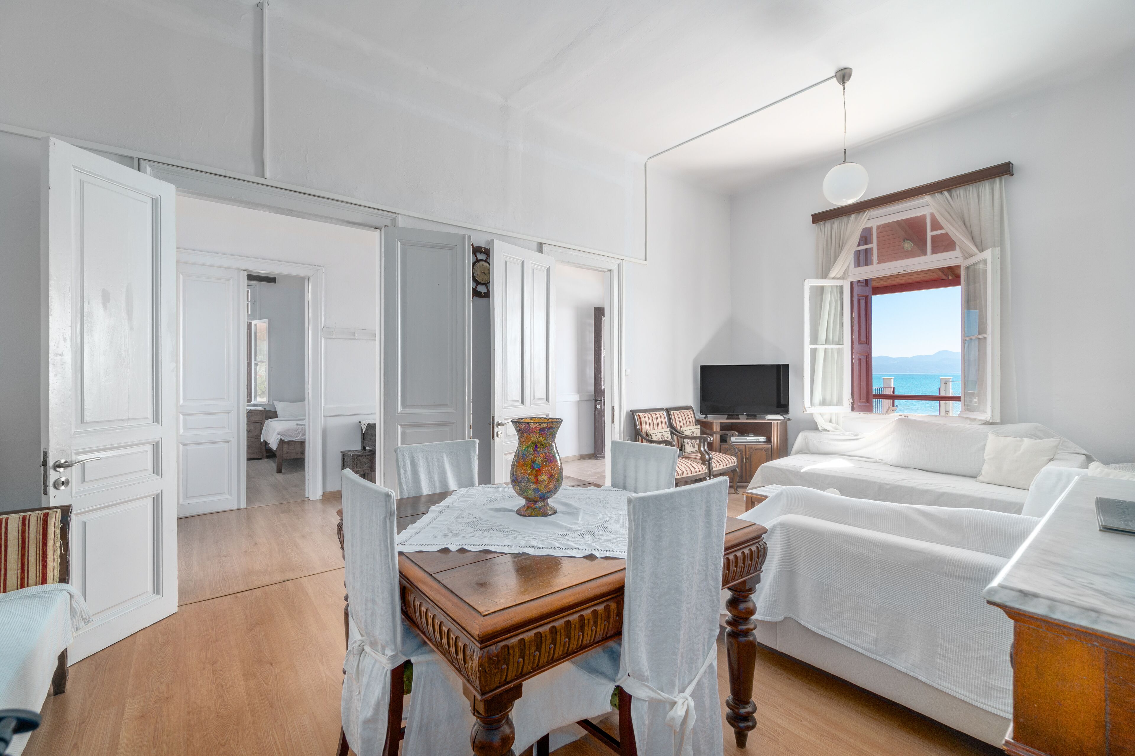 Ampla sala de estar com uma mesa de jantar de madeira, mobiliário branco estofado e vista para o mar através de uma janela com cortinas.