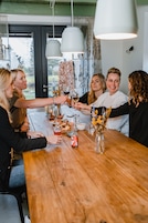 Um grupo de pessoas brindando em uma mesa de madeira, com taças de vinho, velas e um vaso de flores.