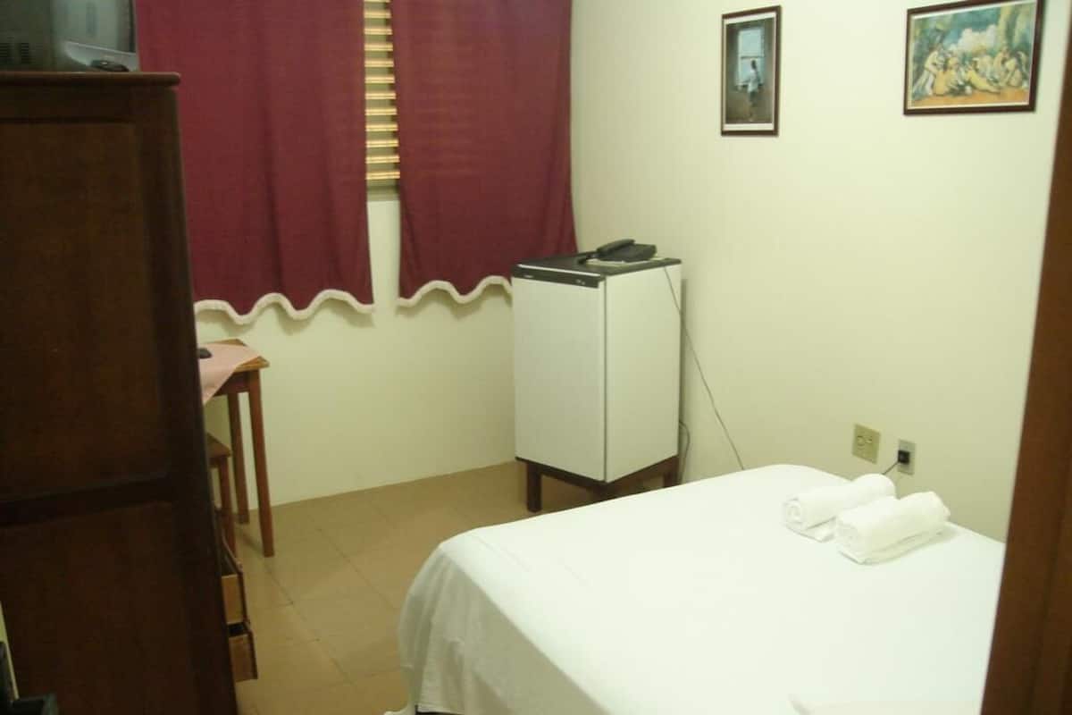 Quarto pequeno e arrumado, com cama, escrivaninha, cadeira, um refrigerador compacto e janela com cortinas.