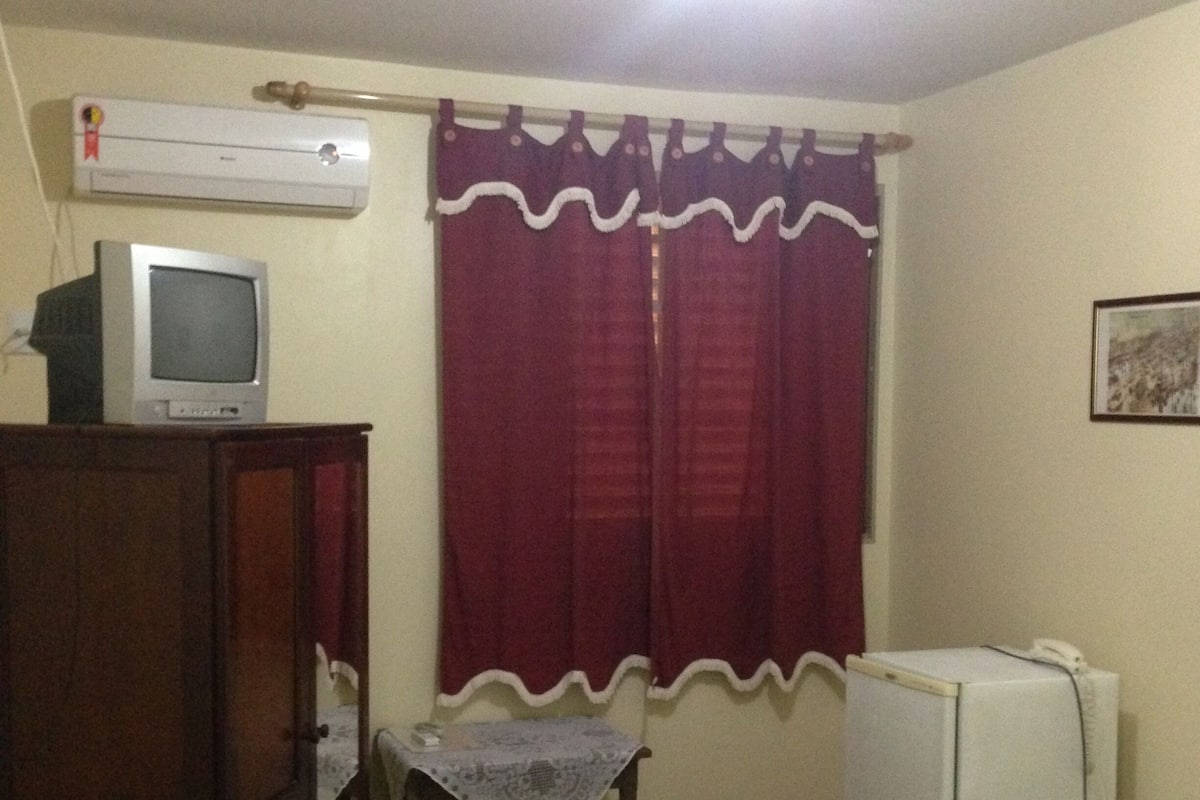 Quarto pequeno com uma cama, uma mesa com toalha, televisão em um móvel, geladeira e janela com cortinas.