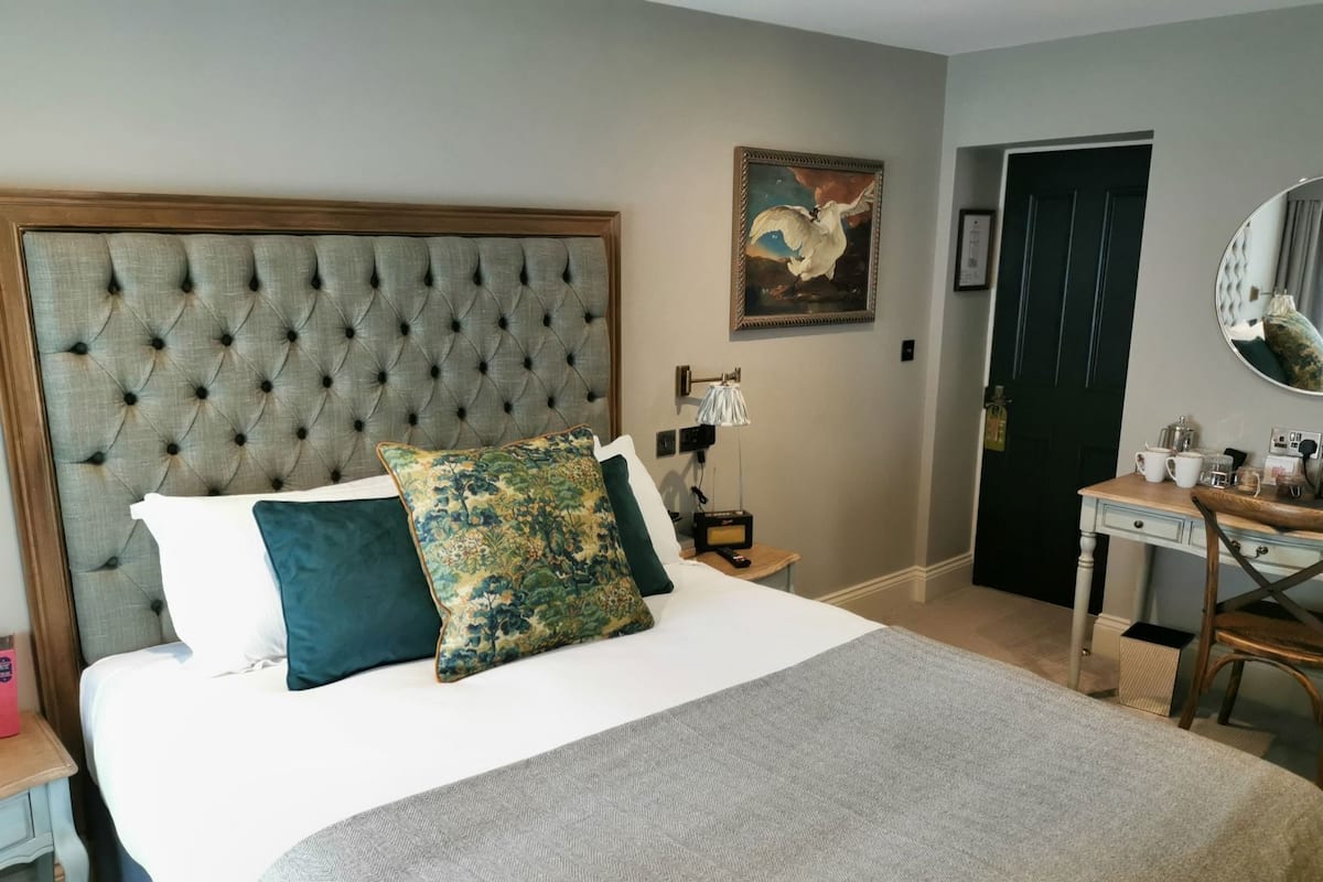 Quarto de hotel com cabeceira estofada, cama com roupa de cama branca e almofadas estampadas, escrivaninha com abajur, quadro na parede e uma porta.