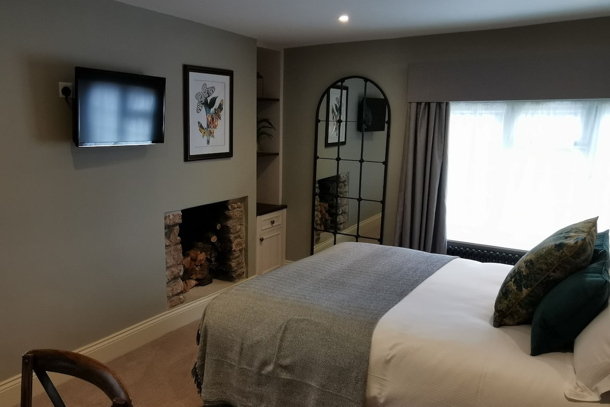 Quarto com cama, televisão fixada na parede, lareira e janela com cortinas.
