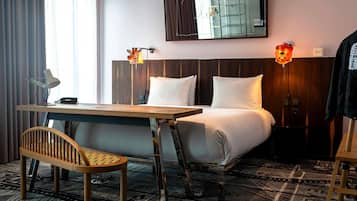 Quarto de hotel com escrivaninha de madeira, uma cadeira, cama com roupa branca, duas luminárias fixadas na parede e um espelho grande refletindo o quarto.