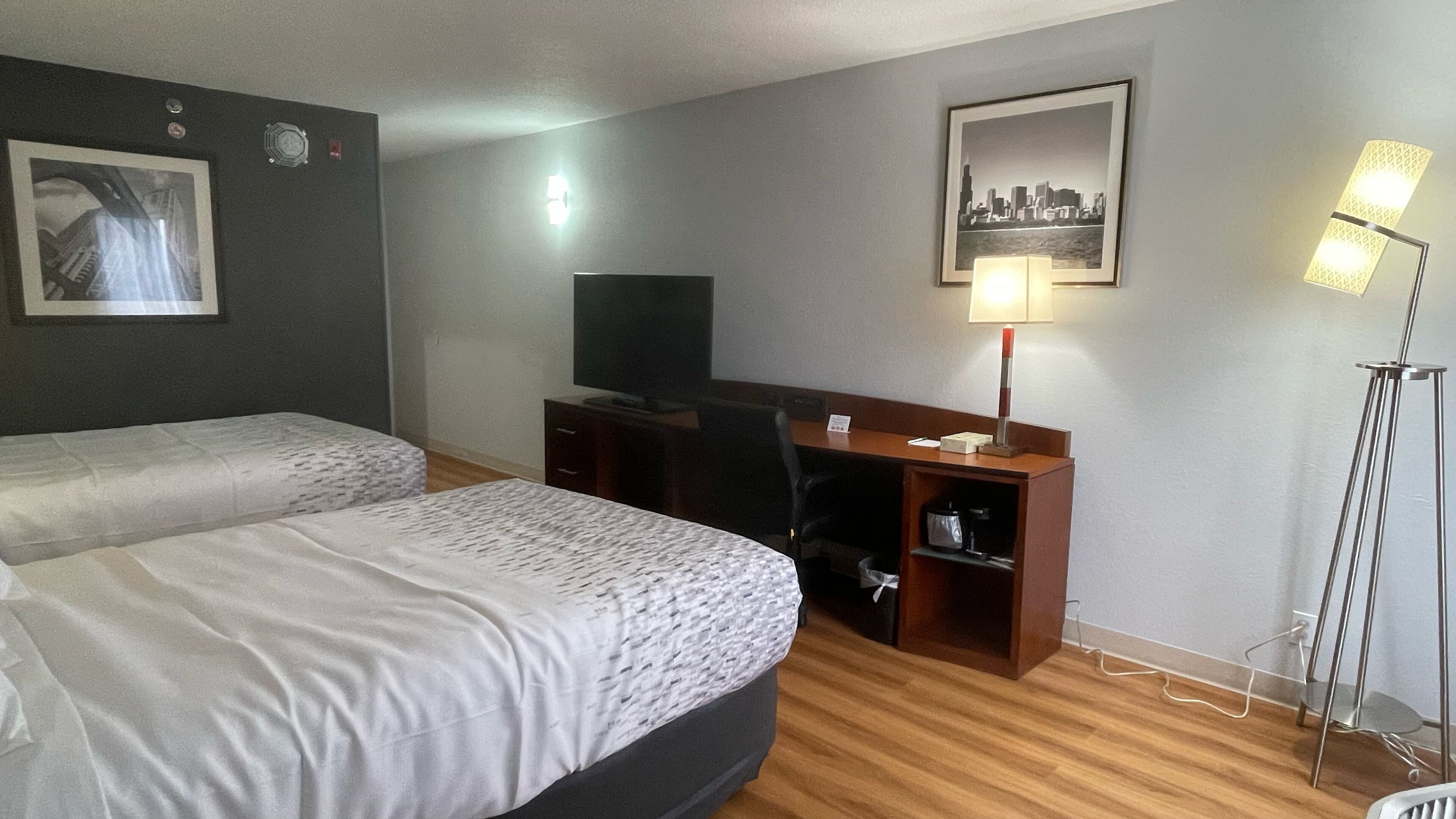Quarto de hotel com duas camas, uma escrivaninha com computador, televisão, abajur e um quadro na parede.