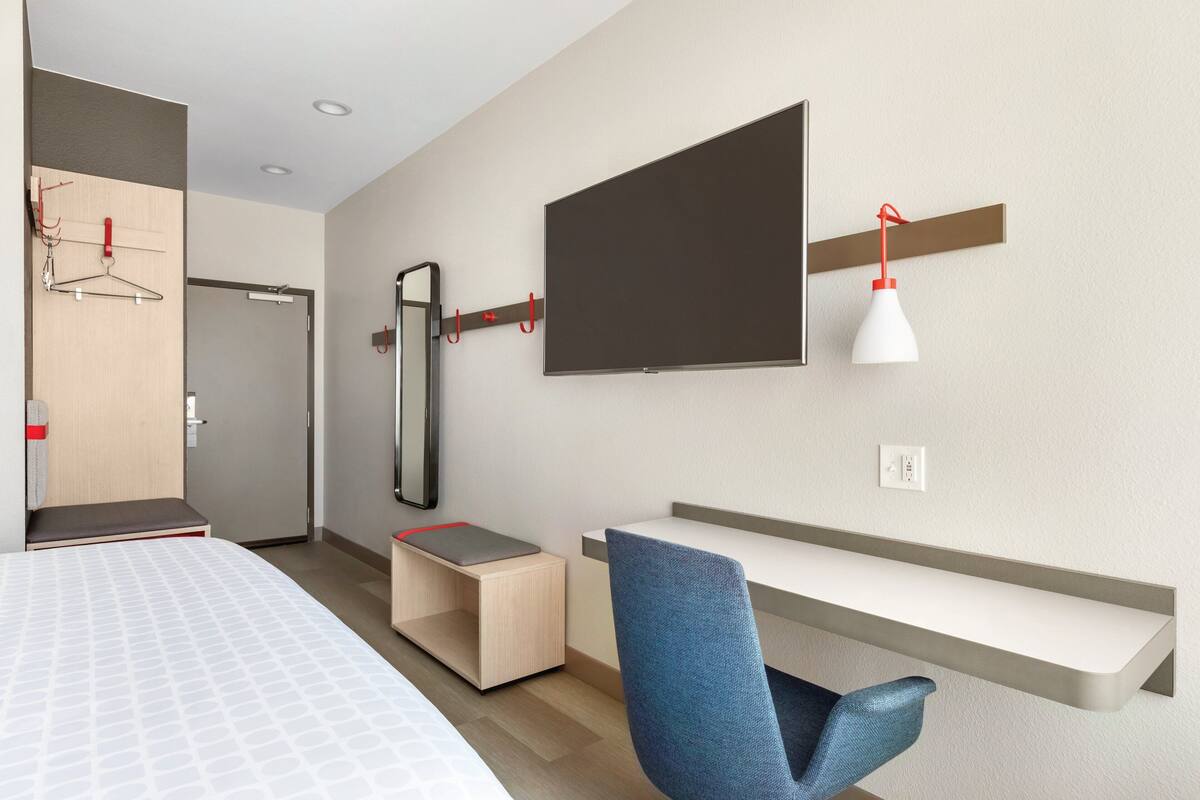 Quarto de hotel com uma cama, escrivaninha, cadeira, televisão e espelho.