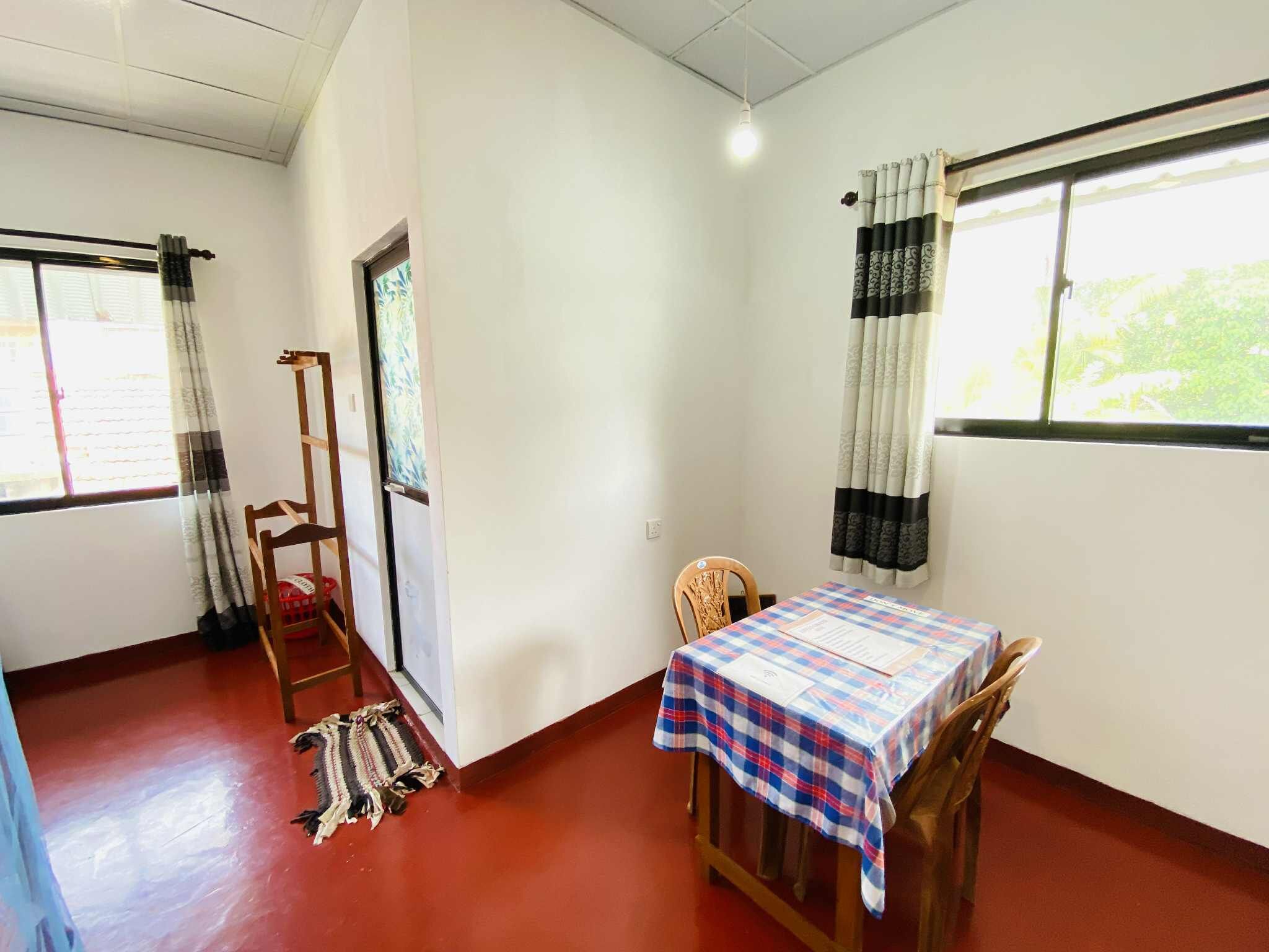 Um quarto com chão vermelho, uma mesa com toalha xadrez, uma cadeira, uma escada de madeira e uma janela com cortinas.
