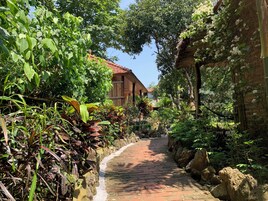Um caminho de tijolos estreito, ladeado por vegetação exuberante e um edifício rústico com telhado de telhas vermelhas.