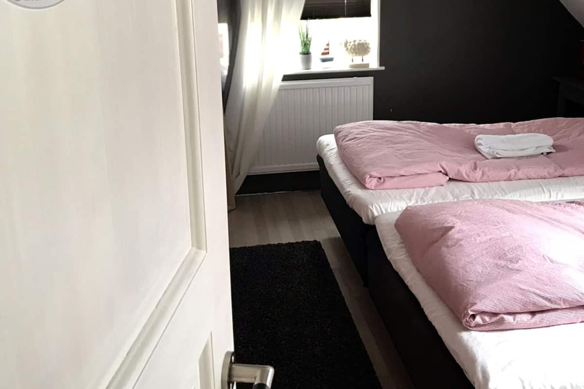 Quarto com cama, janela com cortinas, radiador e porta com aviso.