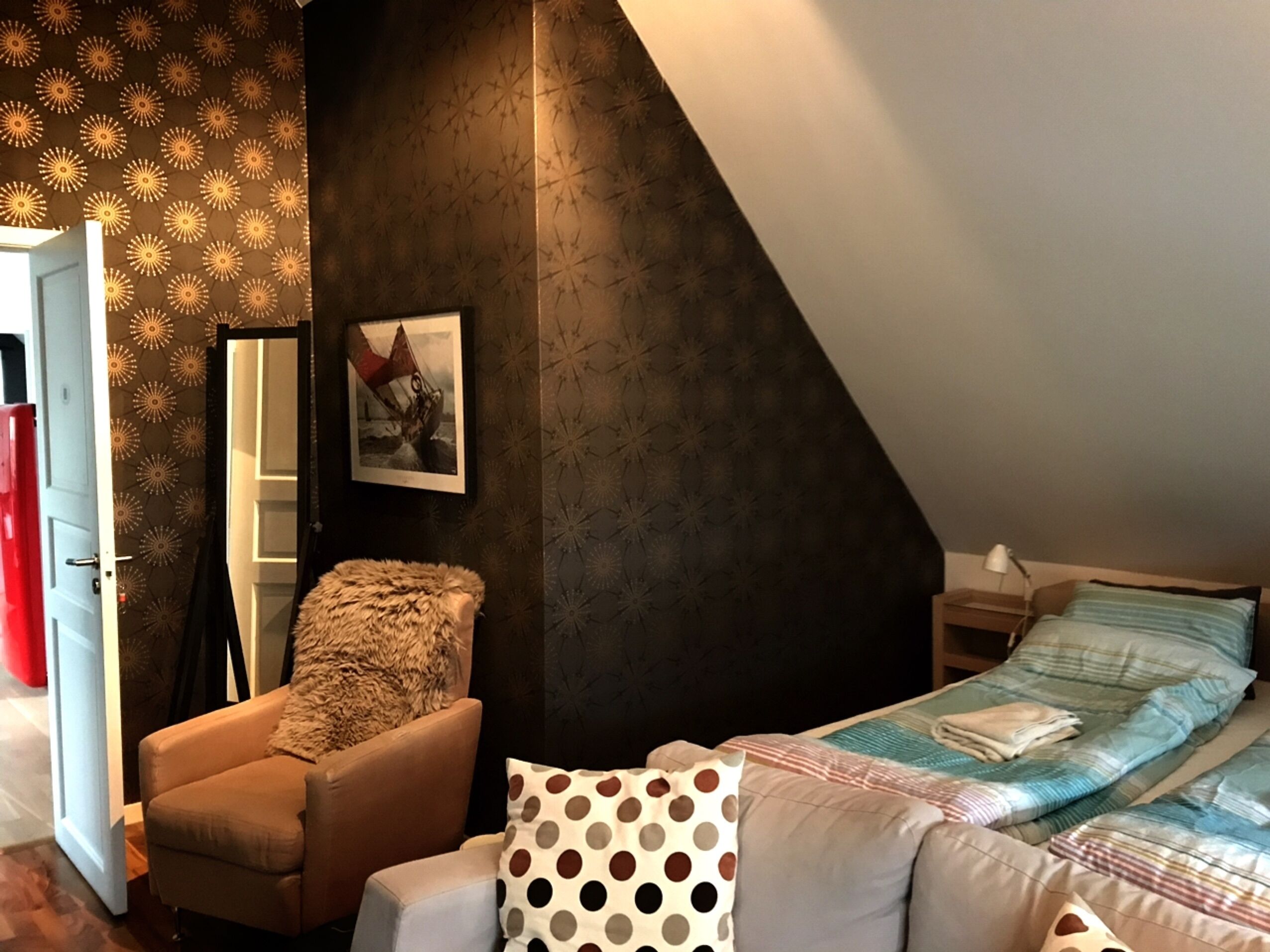 Quarto com teto inclinado, cama com roupa de cama listrada, uma cadeira, um banquinho fofo, um quadro na parede e uma porta com espelho.