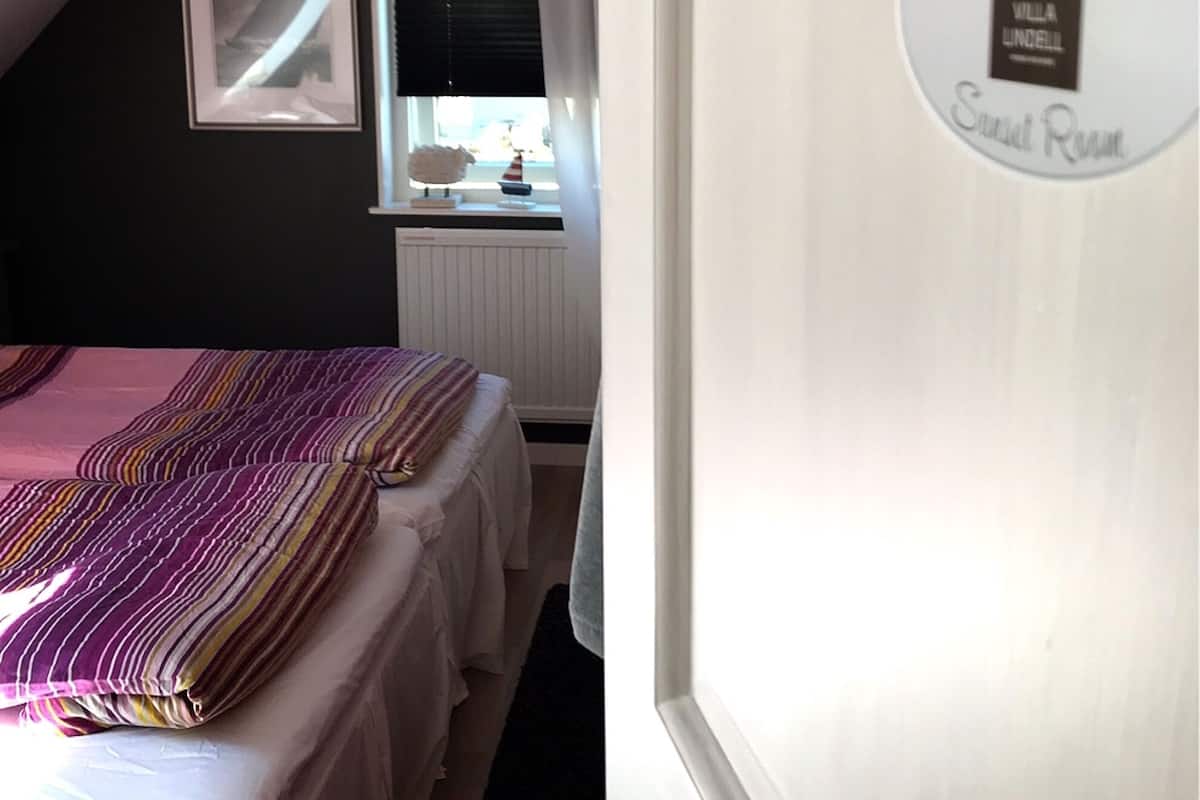 Um quarto com uma cama, janela com cortinas, radiador e um aviso de boas-vindas na porta.
