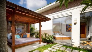 Uma área de estar moderna ao ar livre, com um pérgola de madeira, um banco acolchoado e um pequeno jardim com pedras no chão.