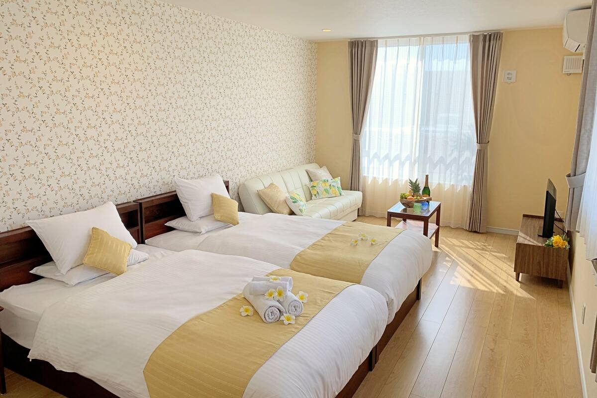 Quarto de hotel com duas camas, um sofá, uma mesinha com uma garrafa e frutas, e uma TV sobre um suporte.