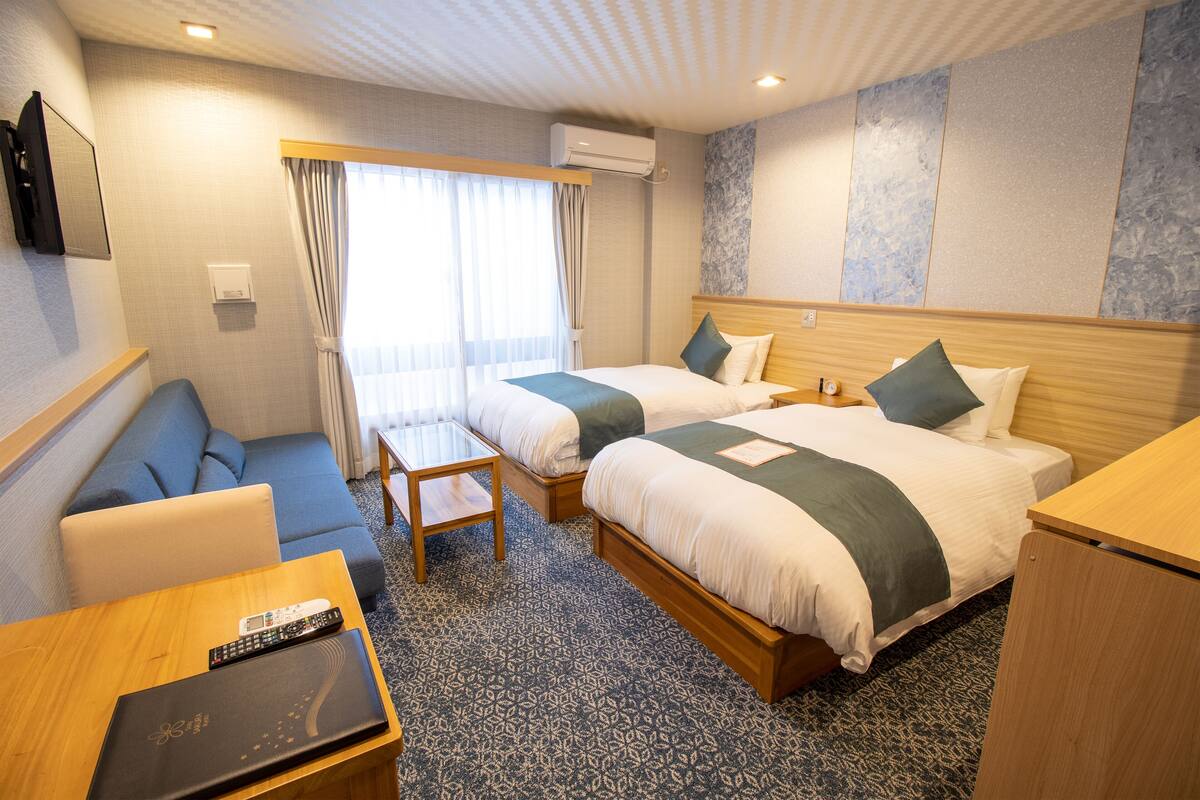 Quarto de hotel com duas camas, um sofá, uma mesinha e uma televisão.
