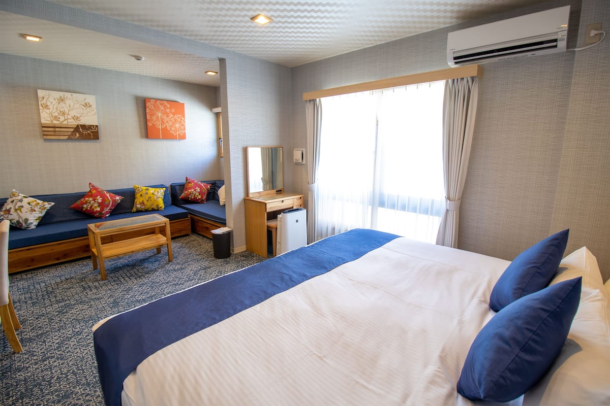 Quarto de hotel com uma cama grande, um sofá com almofadas coloridas, uma mesinha, uma escrivaninha e uma janela com cortinas.