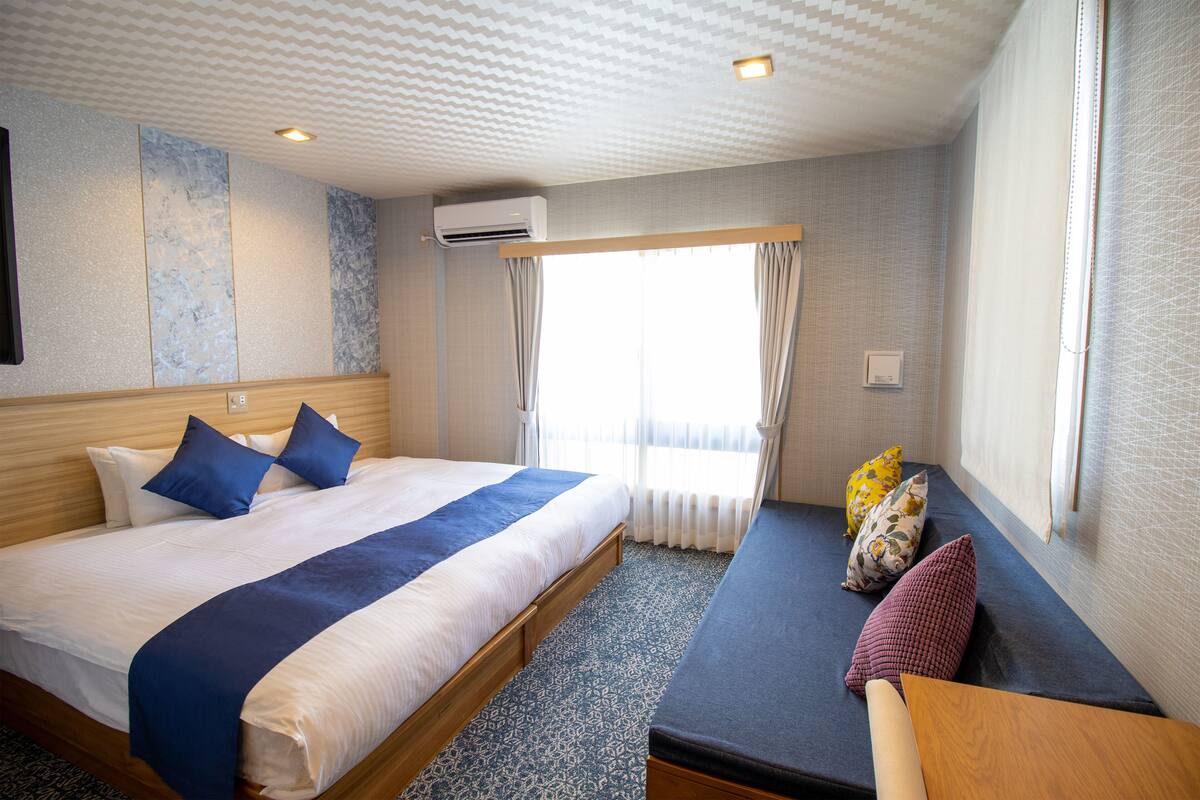 Quarto de hotel com cama, um banquinho e janela com cortinas.